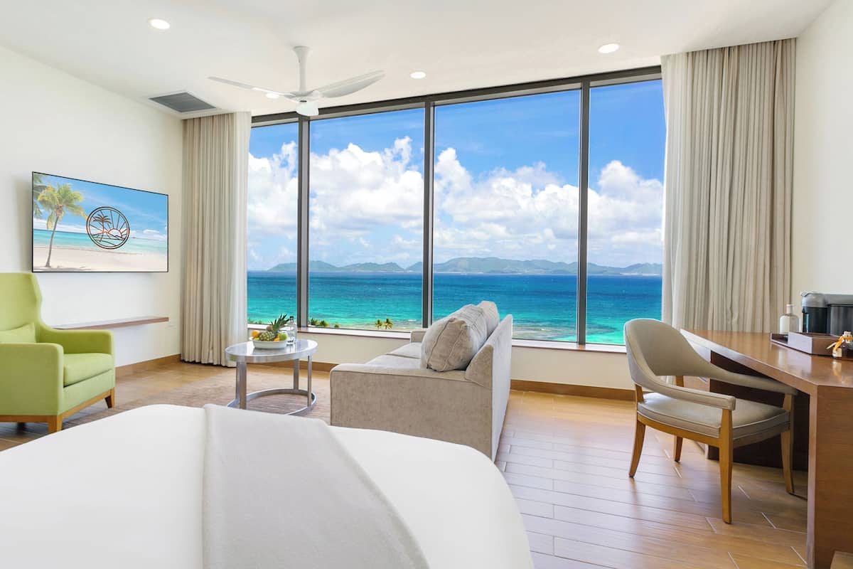 Quarto de hotel com uma grande janela que oferece vista para o oceano e montanhas. Há uma cama, uma poltrona verde, um sofá e uma mesa com cadeira.