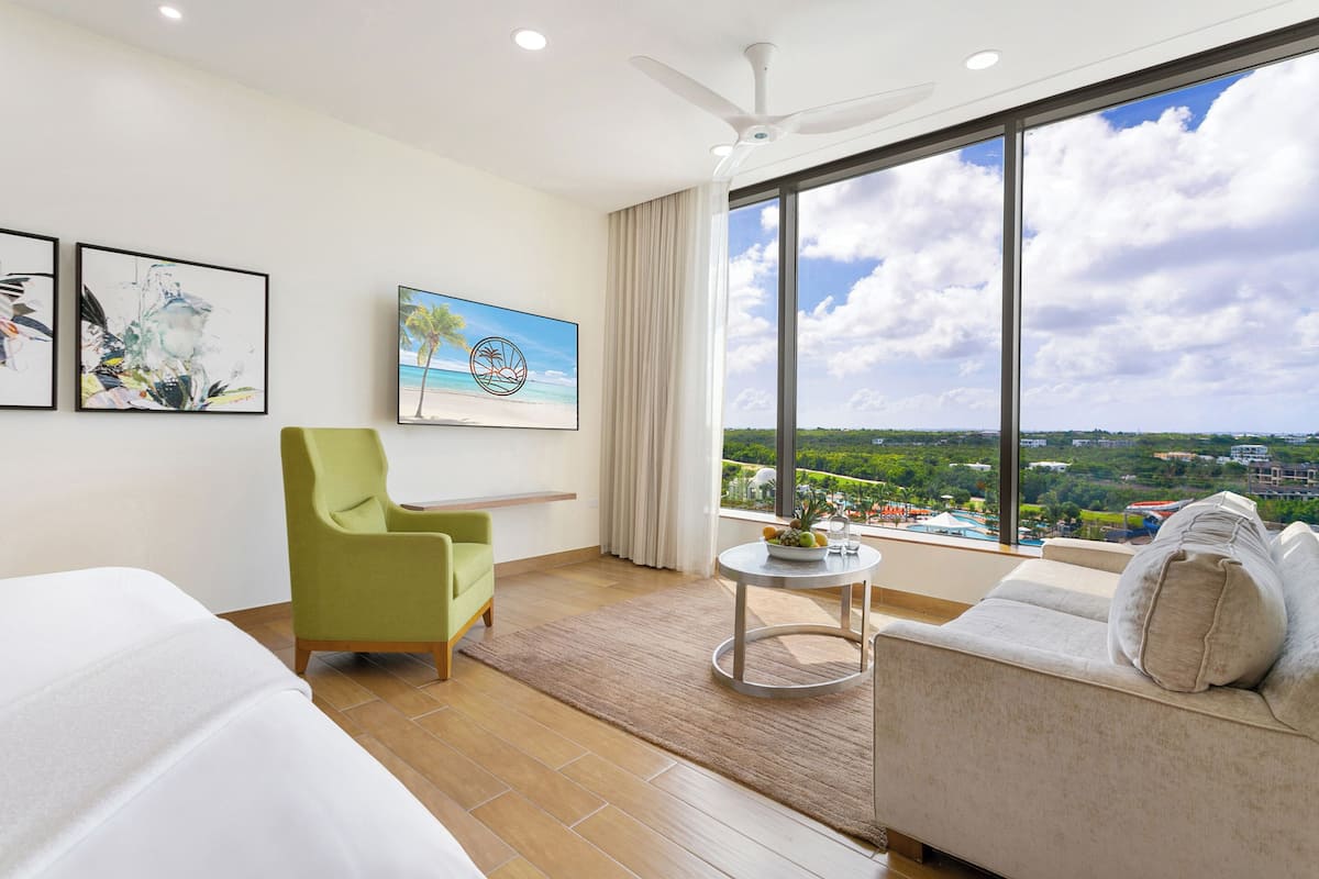 Quarto de hotel moderno com uma janela grande, uma poltrona verde, um sofá e obras de arte nas paredes.