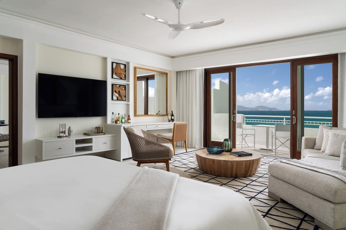 Quarto de hotel com uma cama grande, uma escrivaninha com televisão de tela plana, uma cadeira, uma mesa de centro redonda e uma varanda com vista para o mar.