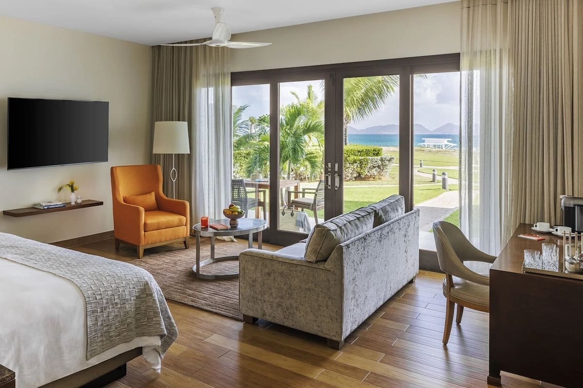Quarto de hotel com cama, sofá, poltrona, televisão, mesa com uma tigela de frutas e vista para o exterior.