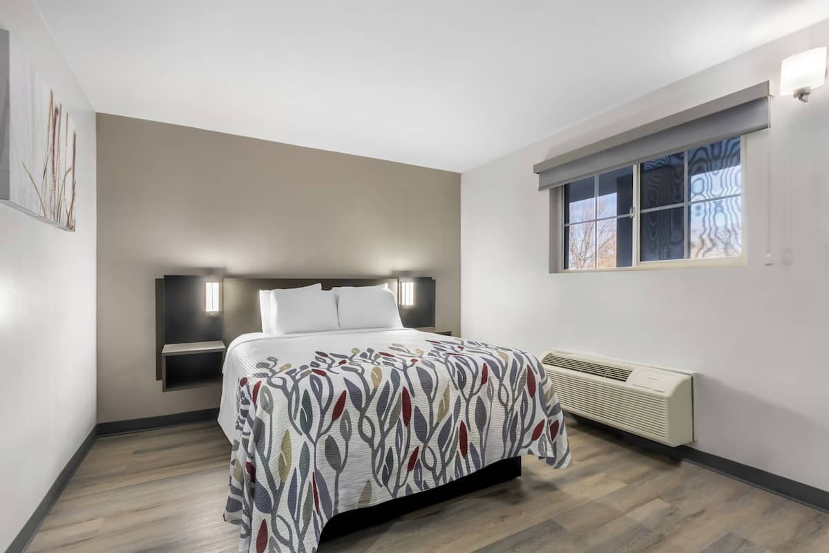 Quarto com cama, mesas de cabeceira, janela com persianas estampadas e ar condicionado fixado na parede.
