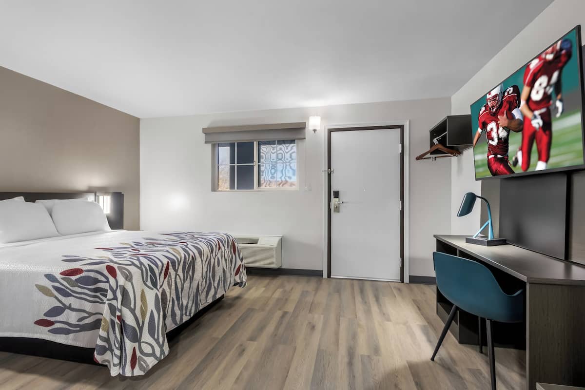 Quarto de hotel moderno com cama, escrivaninha, cadeira e televisão exibindo uma partida de futebol.