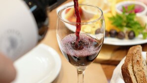 Uma taça de vinho sendo servida com vinho tinto, um prato de queijo e pão sobre uma mesa de madeira.