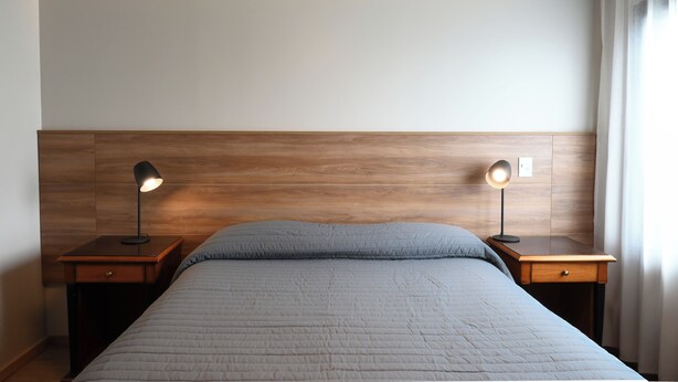 Quarto com cabeceira de madeira, cama com colcha cinza, duas mesas de cabeceira com luminárias e uma janela com cortinas.
