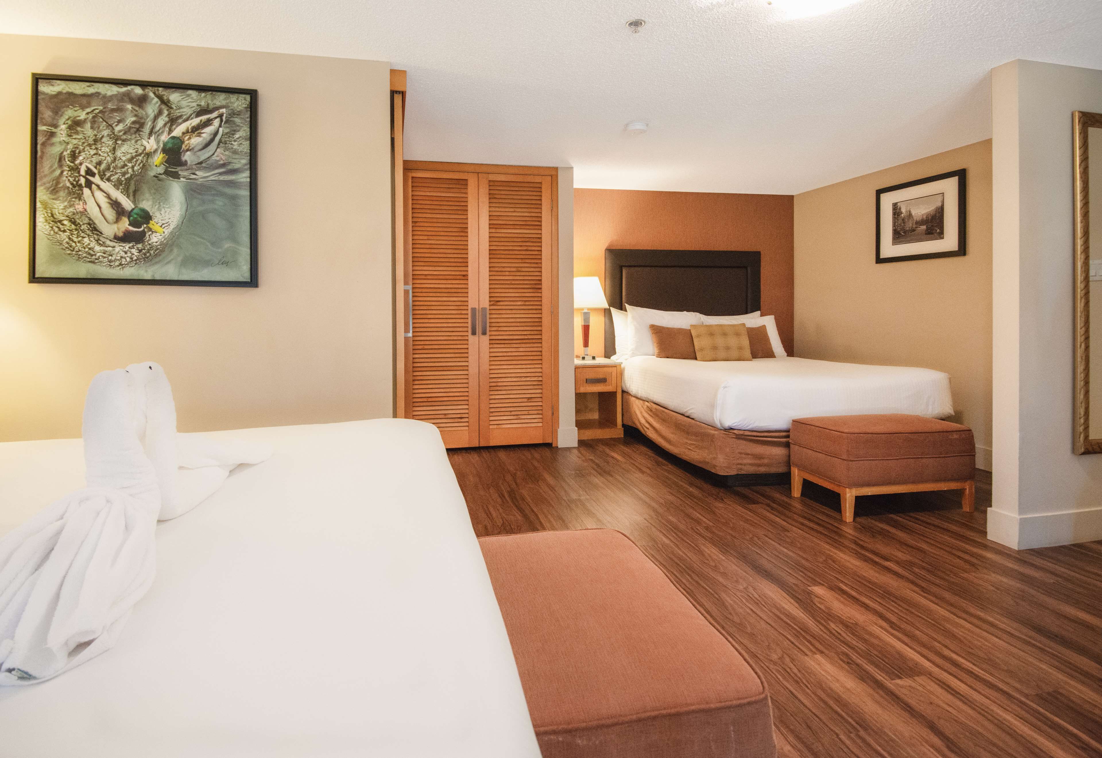 Quarto de hotel com uma cama grande, uma cama menor, um guarda-roupa de madeira, uma mesa de cabeceira com abajur e uma moldura com quadro na parede.