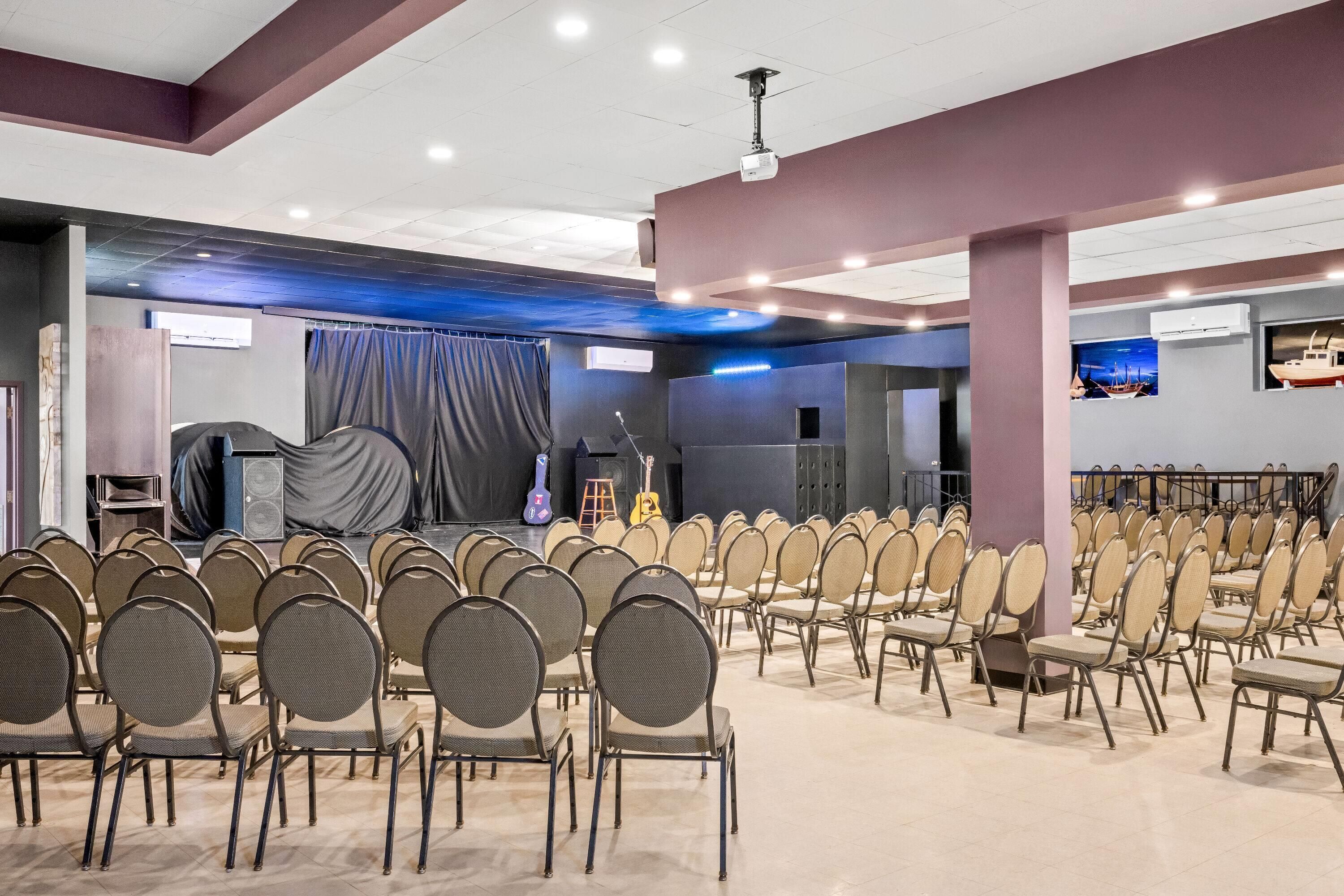 Uma sala de conferências com fileiras de cadeiras, um palco com instrumentos musicais e um projetor.