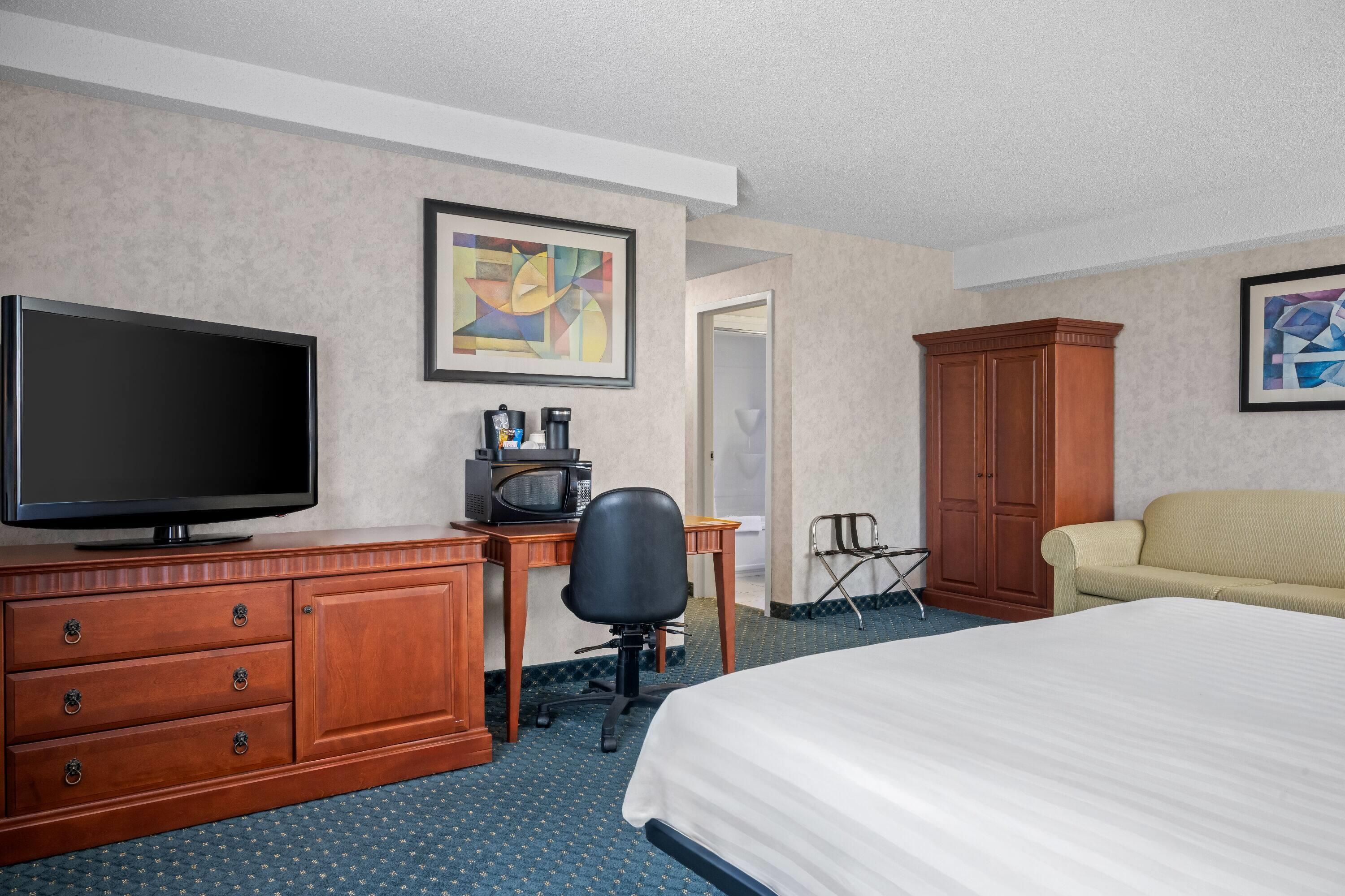 Quarto de hotel com cama, televisão, escrivaninha com cadeira, guarda-roupa e duas pinturas na parede.