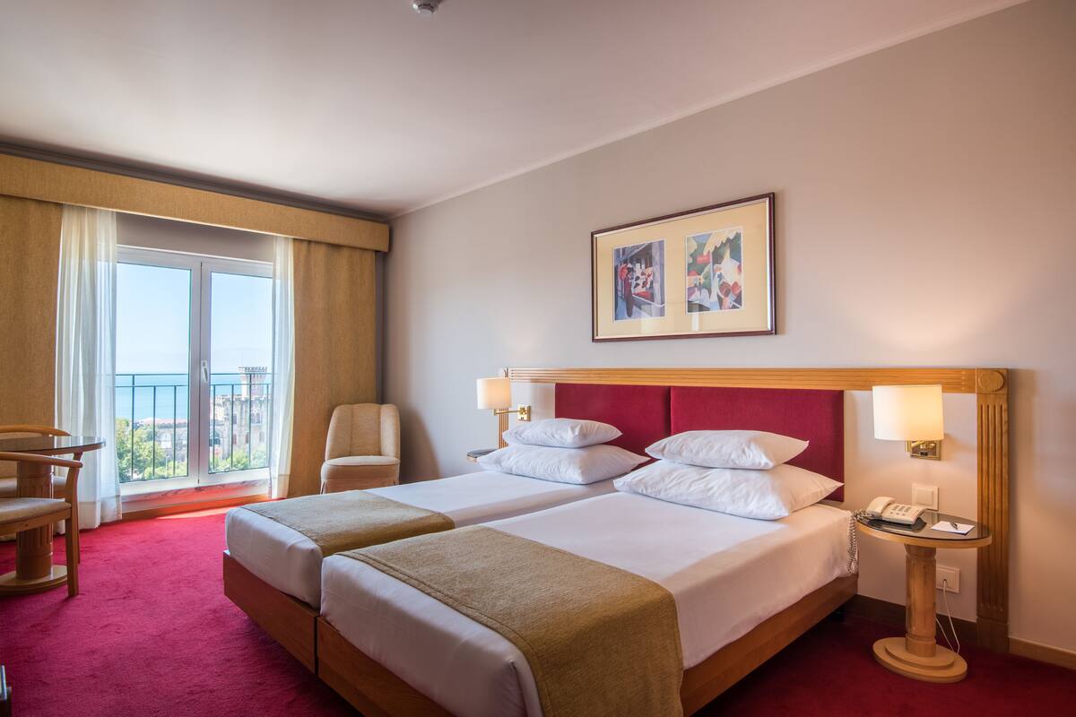 Quarto de hotel com duas camas, uma escrivaninha, uma cadeira, uma varanda e um quadro na parede.
