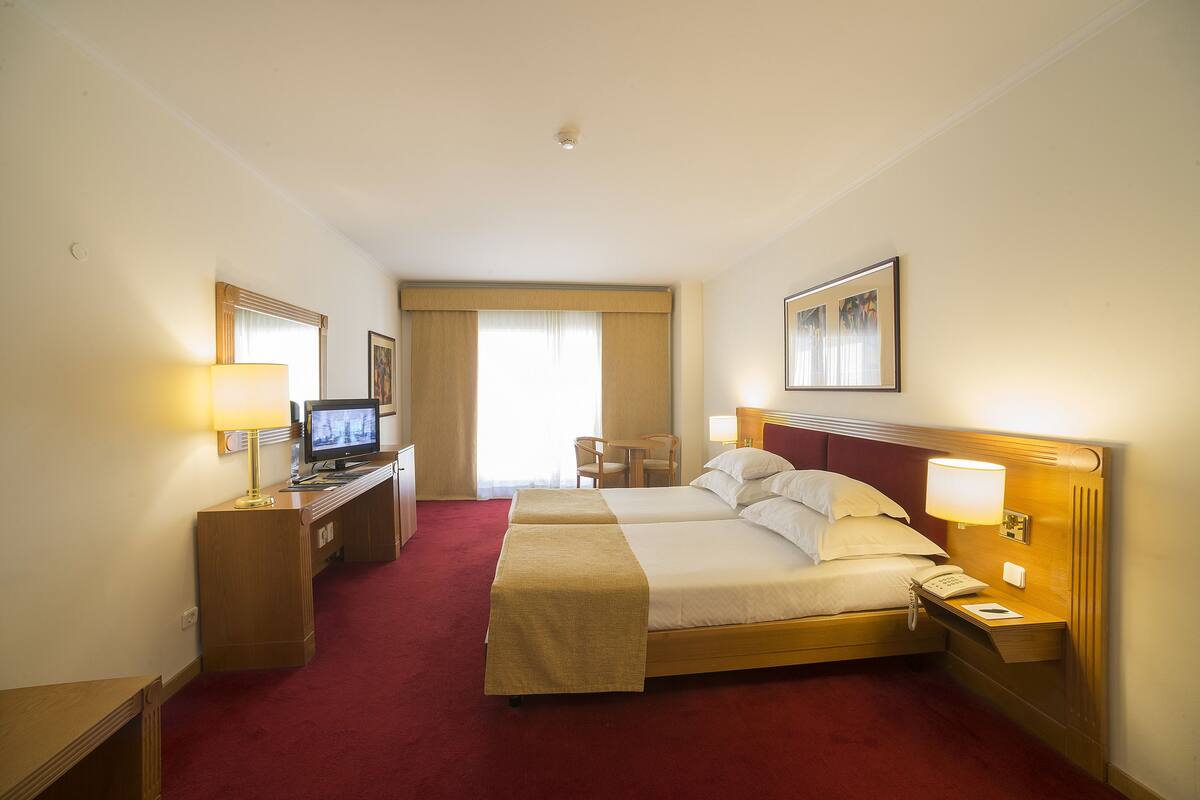 Quarto de hotel com duas camas, uma escrivaninha com televisão, uma cadeira e uma janela com cortinas.