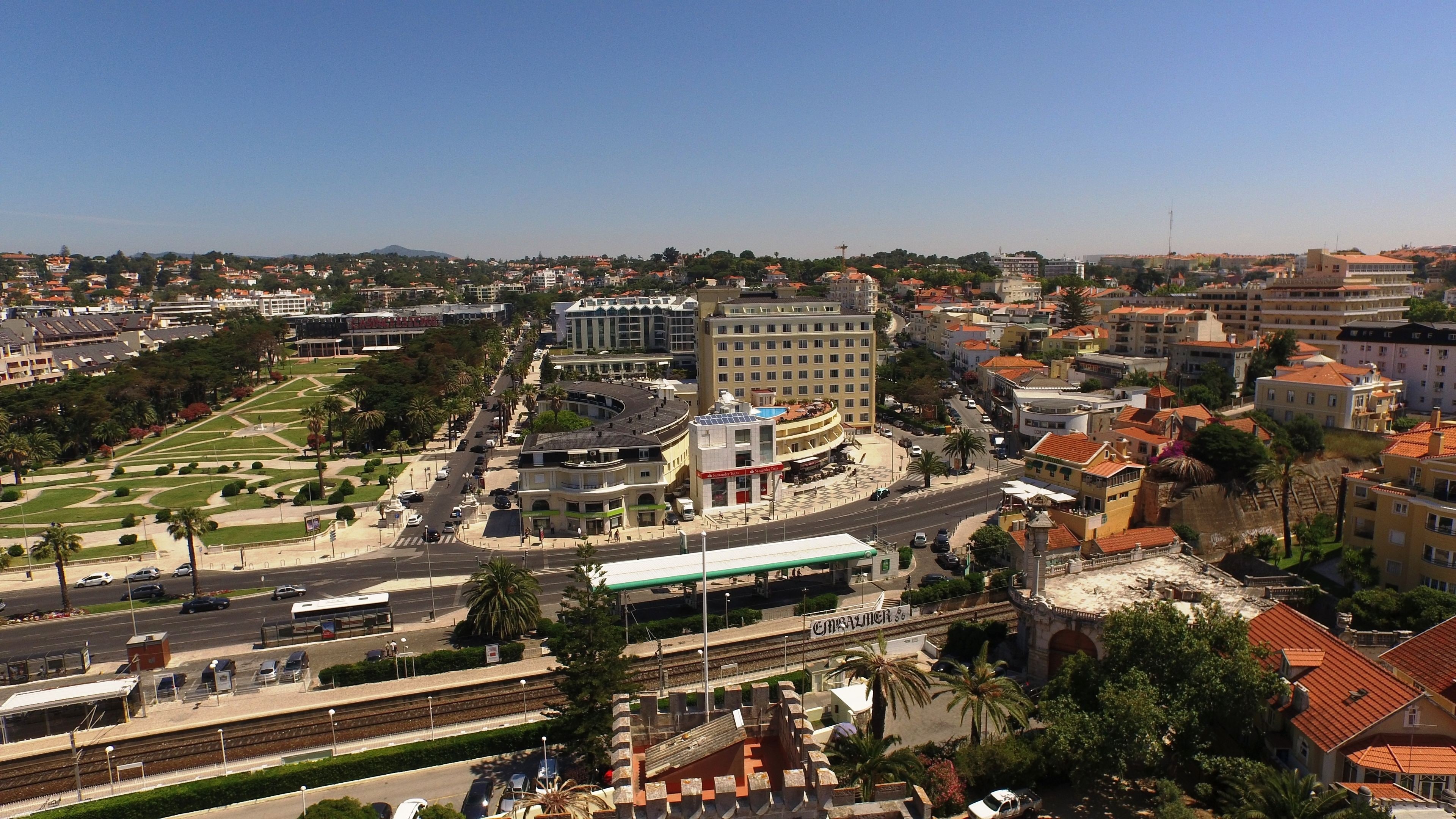 Vista urbana com um edifício circular imponente com cúpula, rodeado por edifícios residenciais e uma praça pública bem cuidada com palmeiras.