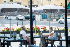 Uma área externa com mesas e cadeiras sob guarda-sóis, com vista para a rua ao fundo.