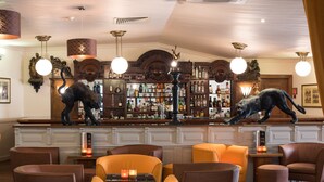 Uma área de bar com balcão de madeira, diversas opções de assento incluindo cadeiras e pufes, e variedade de garrafas e copos em prateleiras.