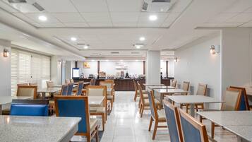 Uma sala de jantar moderna com várias mesas e cadeiras, um balcão central com exposição de menu e um ambiente limpo e bem iluminado.