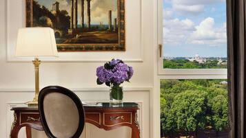 Quarto com escrivaninha, abajur e um vaso de flores. Há um quadro na parede e uma janela oferece vista para árvores e um edifício ao longe.