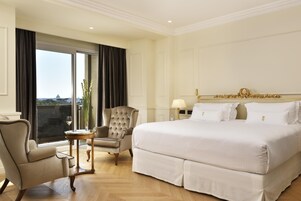 Quarto de hotel com uma cama grande, uma cadeira, uma mesinha e vista para a cidade.