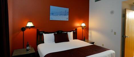 Quarto de hotel com uma cama grande, duas mesas de cabeceira com abajures, cabeceira de madeira, parede vermelha e um quadro de uma paisagem nevada.