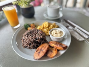 Um prato de arroz preto, banana da terra frita, ovos mexidos e uma pequena tigela de molho sobre uma mesa.
