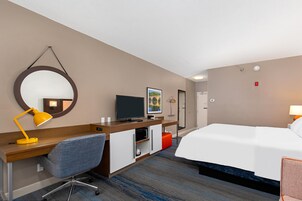 Quarto de hotel com cama, escrivaninha, cadeira, televisão e espelho.