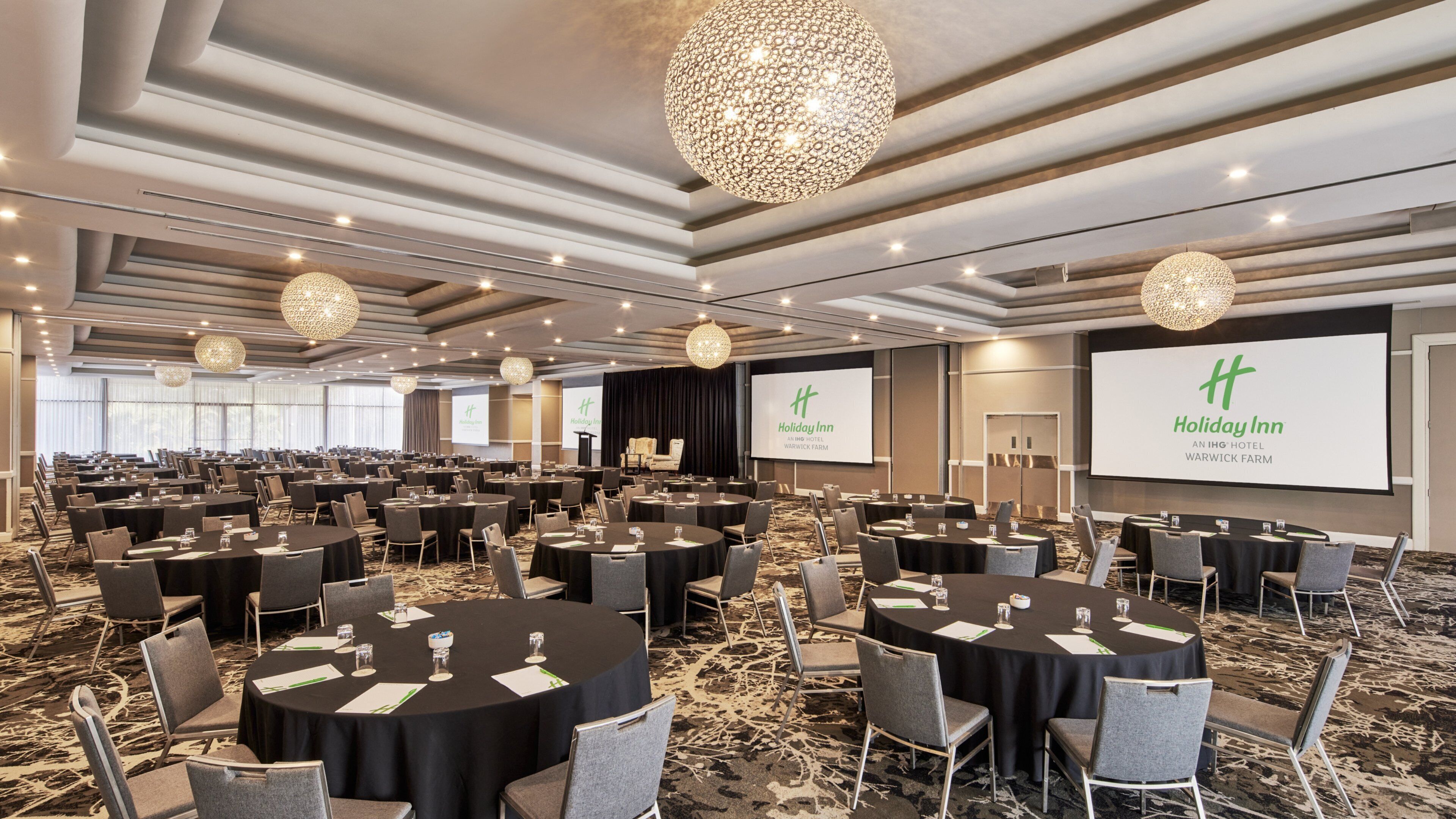 Uma sala de conferências com várias mesas redondas, cadeiras e uma tela grande exibindo o logotipo do Holiday Inn.