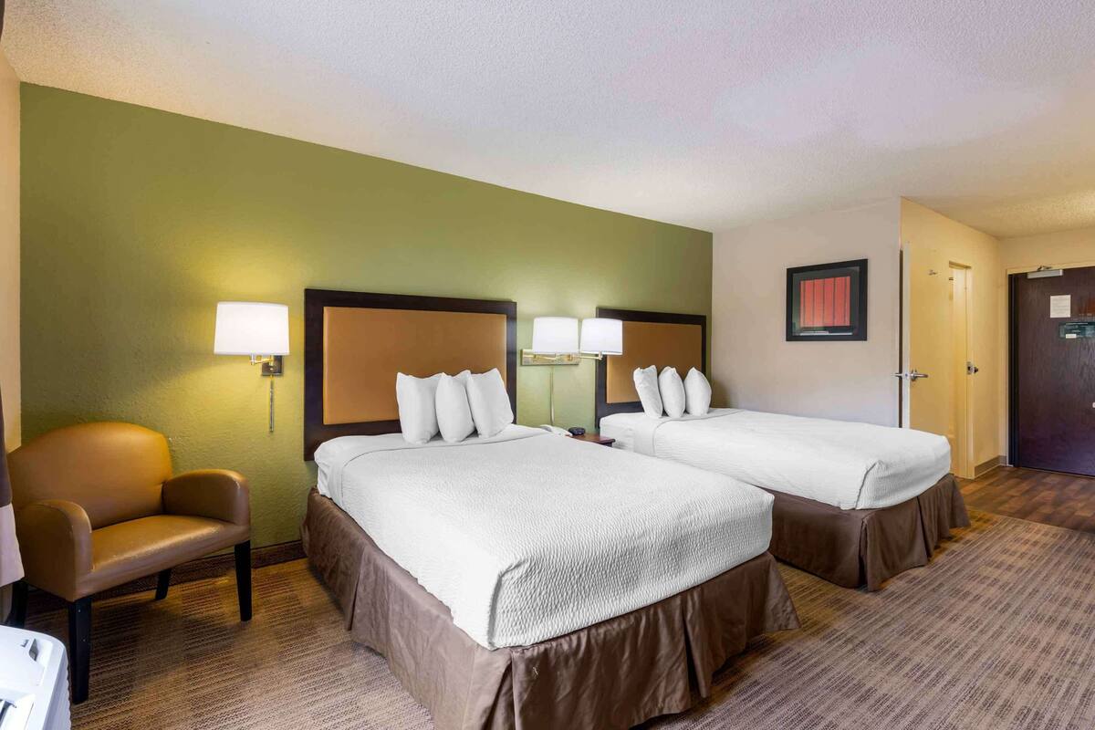 Quarto de hotel com duas camas, uma cadeira, uma escrivaninha e um abajur.