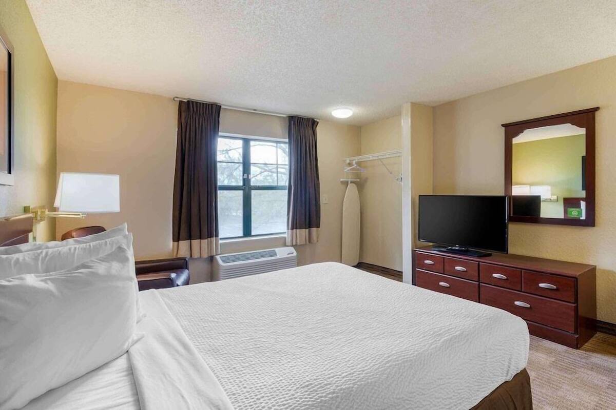 Quarto de hotel com cama, televisão, penteadeira, espelho e janela com cortinas.