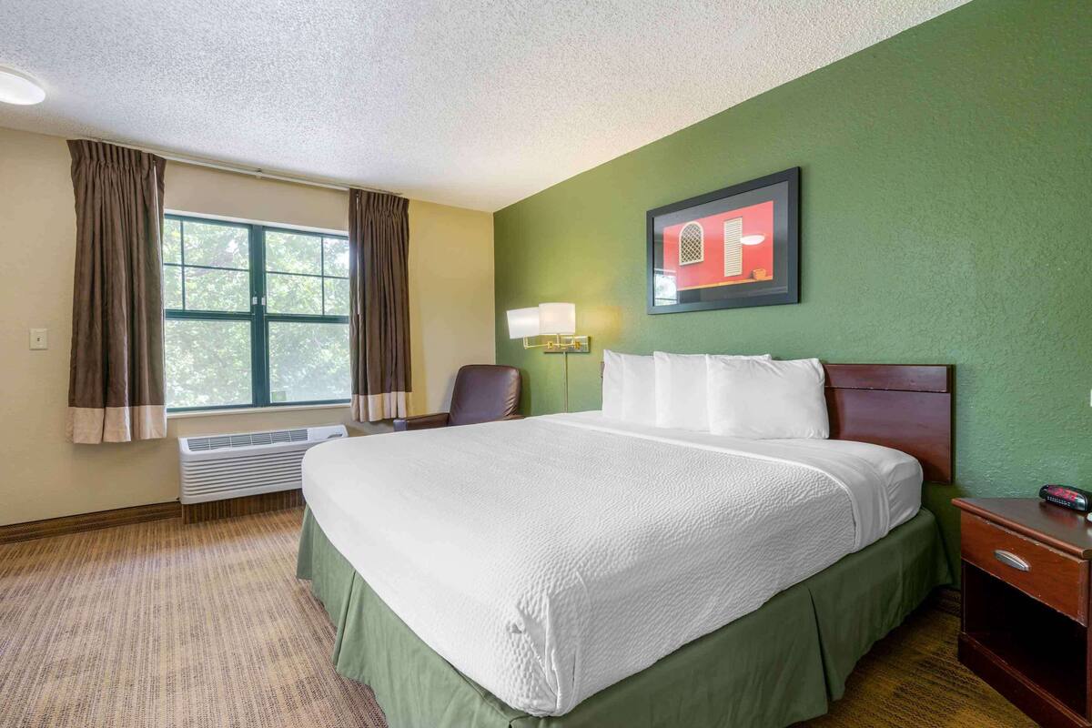 Quarto de hotel com uma cama grande, uma mesa de cabeceira, uma luminária, uma janela com cortinas e um quadro na parede.
