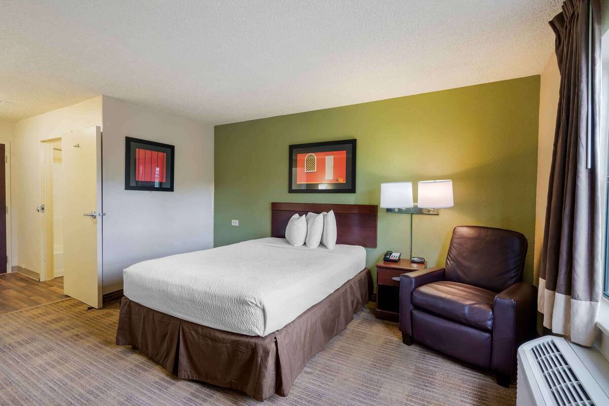 Quarto de hotel com cama, uma cadeira, mesa de cabeceira e janela com cortinas.