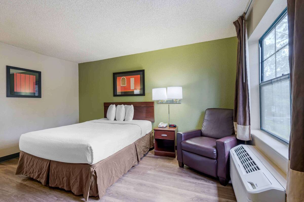 Quarto de hotel com uma cama, uma cadeira, uma mesa de cabeceira com abajur e uma janela com cortinas.