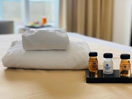 Cama bem arrumada com uma toalha branca dobrada e três frascos de produtos de higiene em um porta-objetos.