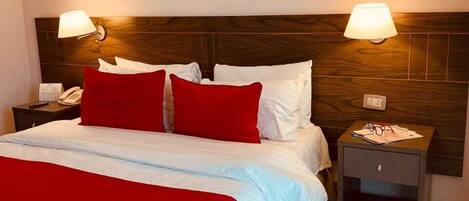 Quarto de hotel com uma cama grande, duas mesas de cabeceira, um criado-mudo e um quadro emoldurado na parede, além de uma luminária em cada mesa de cabeceira.