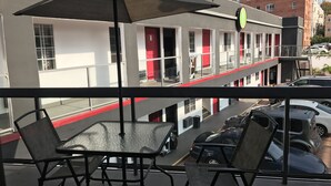 Uma varanda com duas mesas e cadeiras, com vista para um estacionamento e um edifício com portas vermelhas e um letreiro verde.