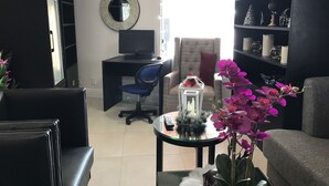 Uma sala de estar com uma cadeira de escritório preta, uma cadeira de escritório azul, uma mesa de centro de vidro com um candeeiro branco e uma estante com objetos decorativos.