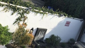 Uma área de estacionamento com uma parede branca, um carro preto e um pequeno jardim com plantas e um lanterna preta.