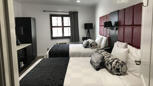 Quarto de hotel com duas camas, uma escrivaninha, um guarda-roupa, uma janela com cortinas e uma televisão.