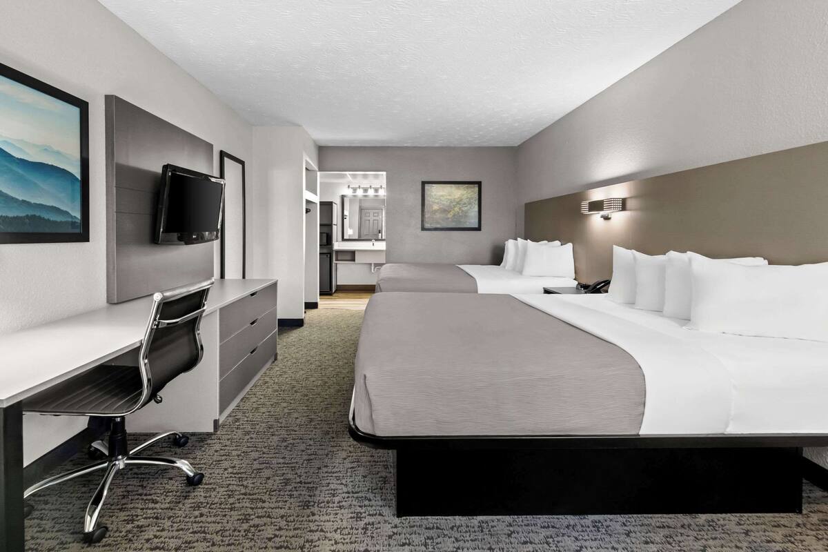 Quarto de hotel com uma cama grande, uma escrivaninha com cadeira, televisão e um banheiro visível ao fundo.