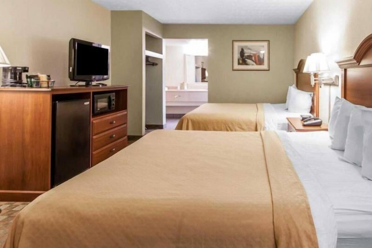 Quarto de hotel com duas camas, uma escrivaninha com televisão, cômoda e banheiro ao fundo.