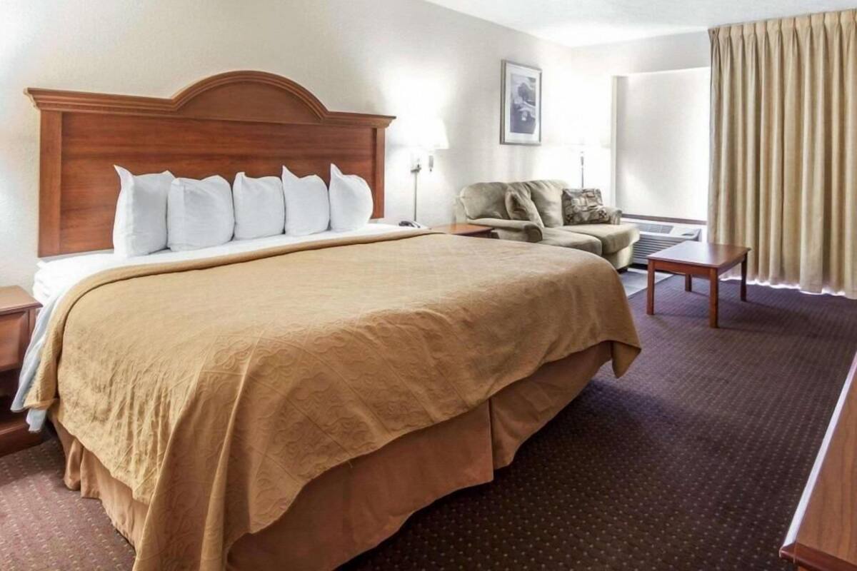 Quarto de hotel com cama grande, cabeceira de madeira, escrivaninha, cadeira e janela com cortinas.