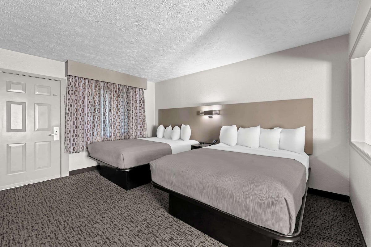 Quarto de hotel com duas camas, cortina estampada, cabeceira com luminária e piso coberto por carpete.