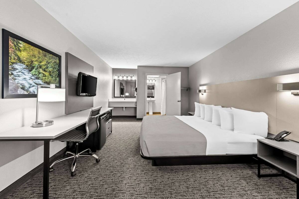 Quarto de hotel com cama, escrivaninha, cadeira, televisão e banheiro visível por uma porta aberta.