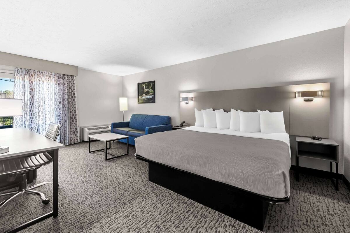 Quarto de hotel com uma cama grande, uma escrivaninha, uma cadeira e uma pequena mesa. Há uma janela com cortinas, uma luminária na parede e um quadro na parede.