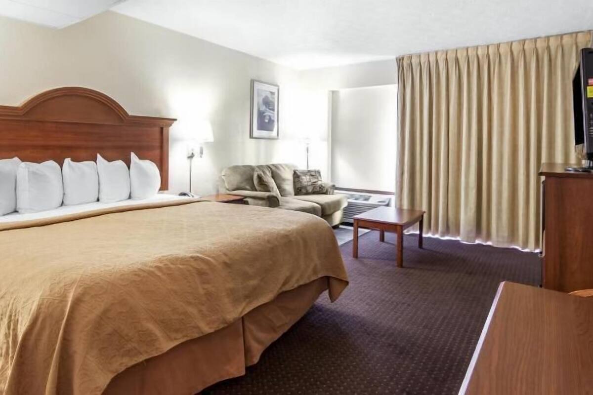 Quarto de hotel com uma cama grande, uma escrivaninha, uma cadeira, uma televisão e uma janela com cortinas.