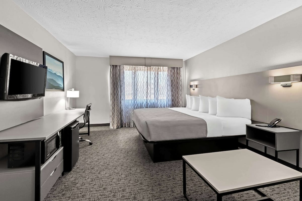 Quarto de hotel com cama, escrivaninha, televisão, luminária e vista para o exterior através das cortinas.