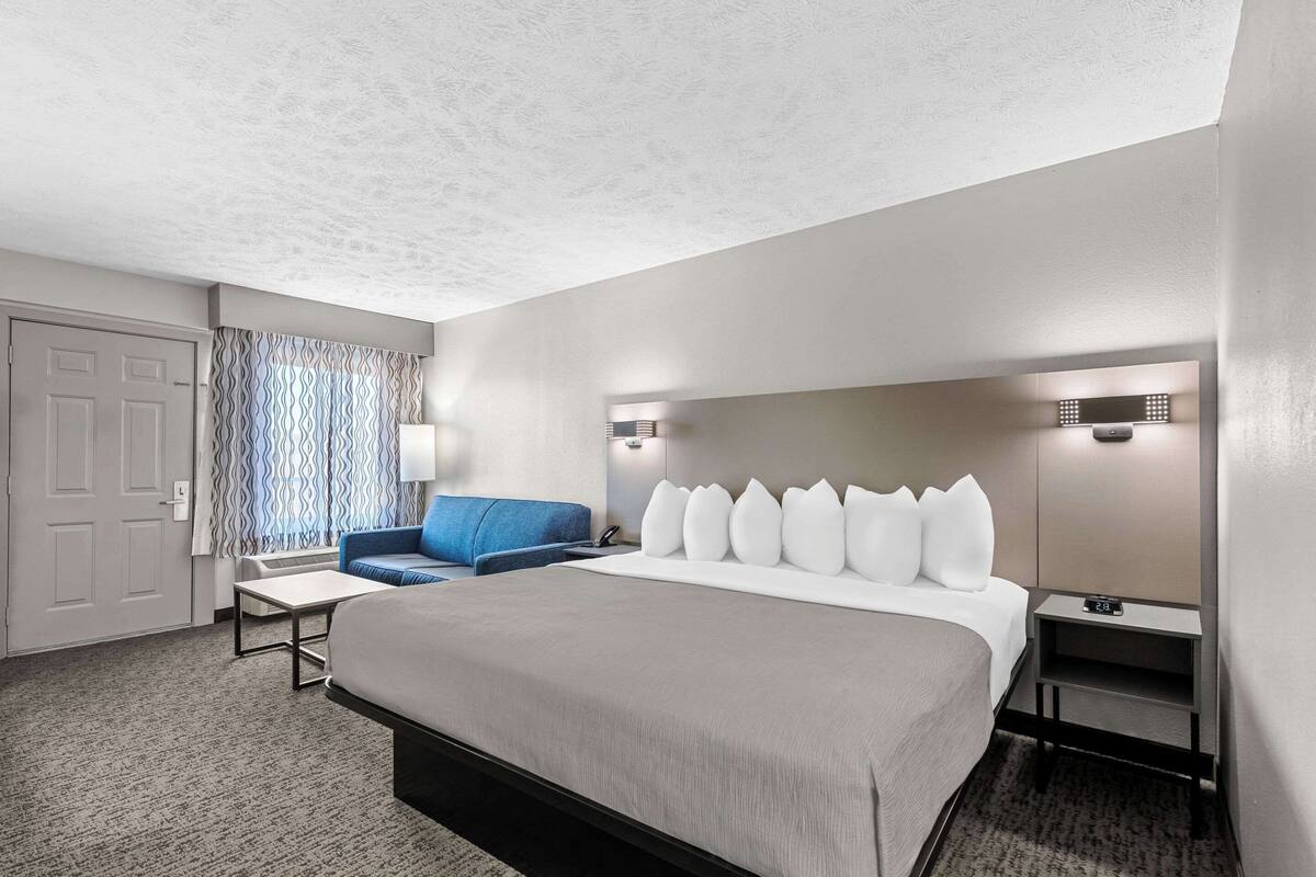 Quarto de hotel com uma cama grande, um sofá azul, uma escrivaninha e uma mesinha de cabeceira com um relógio.