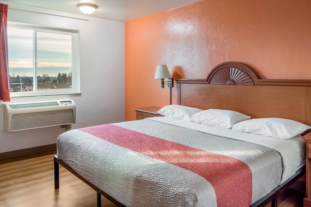 Quarto de hotel com cama, mesa de cabeceira, abajur e janela com cortinas.