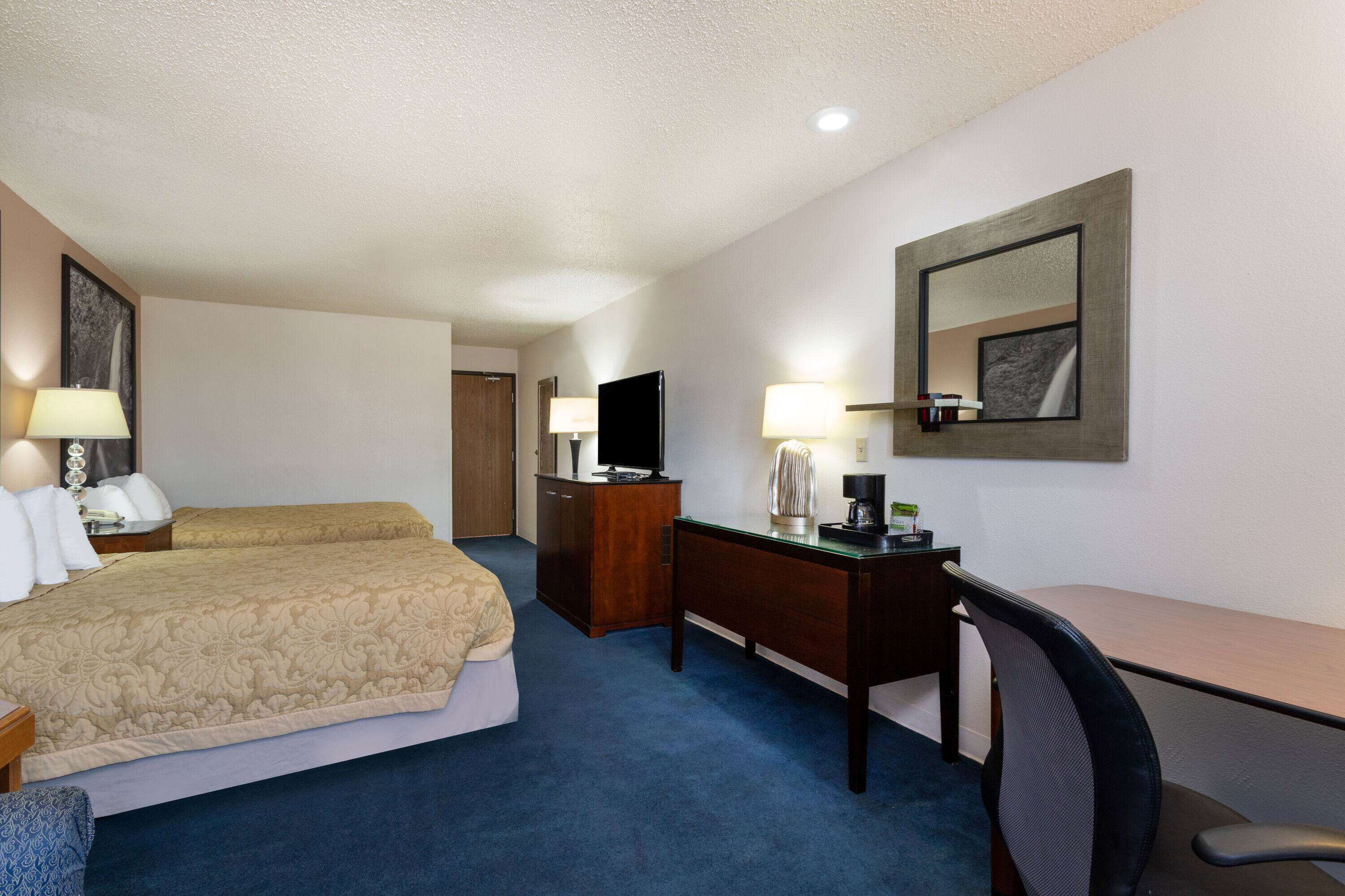Quarto de hotel com duas camas, uma escrivaninha, uma cadeira, uma televisão e um espelho na parede.
