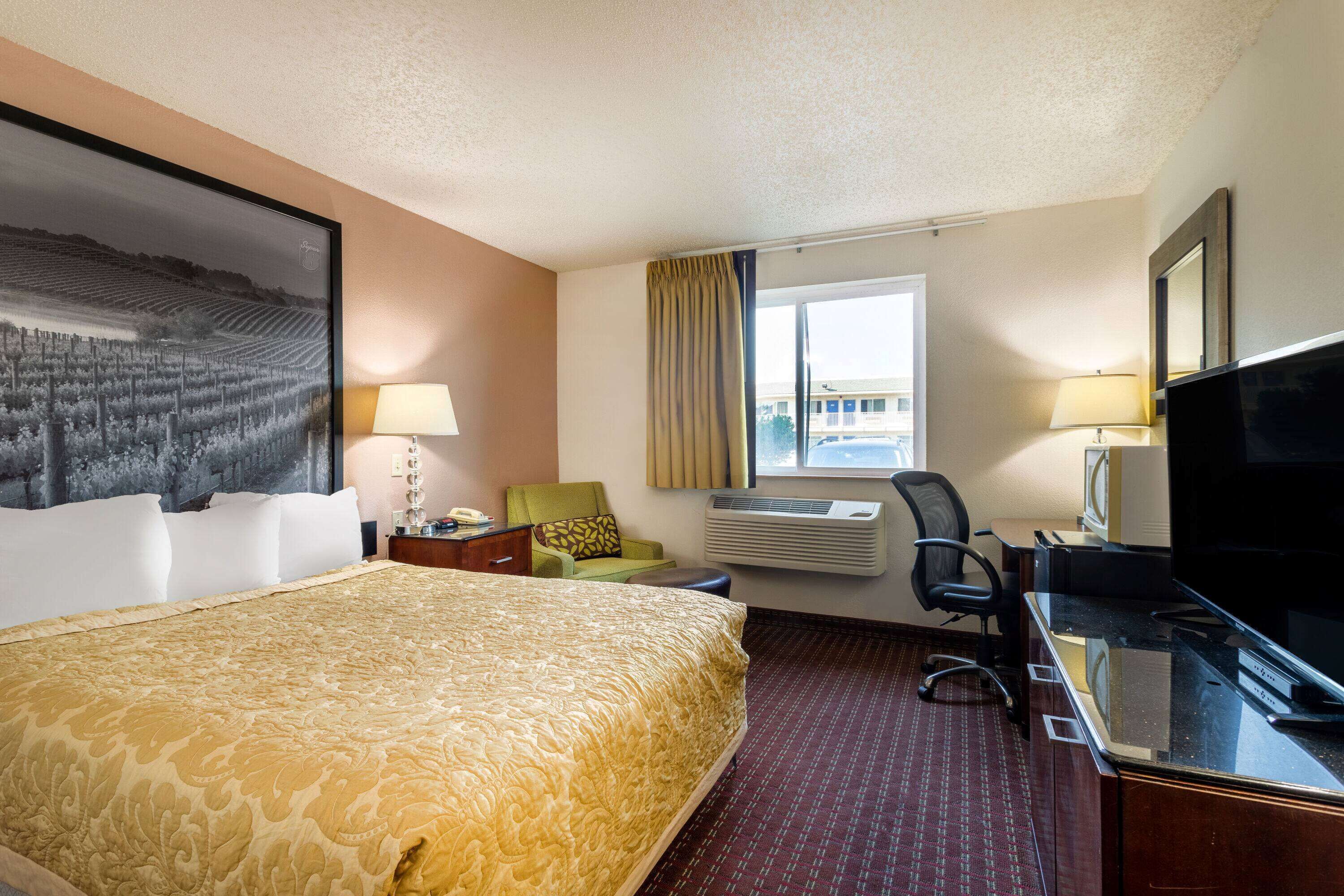 Quarto de hotel com uma cama grande, uma escrivaninha com computador, televisão, ar condicionado e uma janela com cortinas.