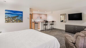 Quarto de hotel com cama, escrivaninha com duas cadeiras, cozinha compacta, televisão e pintura com cena de praia.