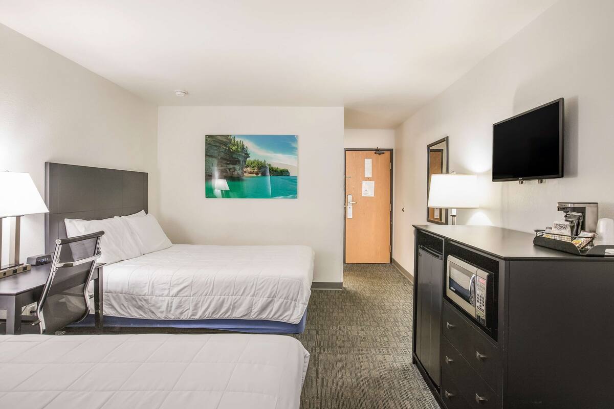 Quarto de hotel com cama, escrivaninha, cadeira, televisão, penteadeira e porta.