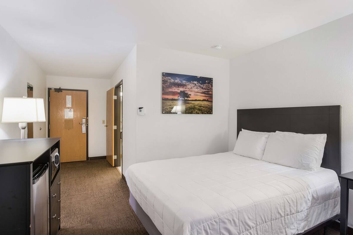 Quarto de hotel com cama, escrivaninha, luminária, porta e quadro fixado na parede.