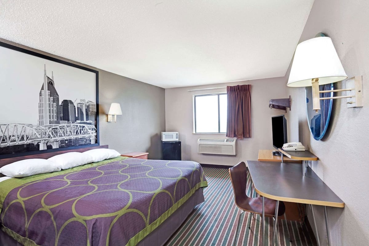 Quarto de hotel com cama, escrivaninha, cadeira, televisão, espelho, janela com cortinas e ar condicionado.